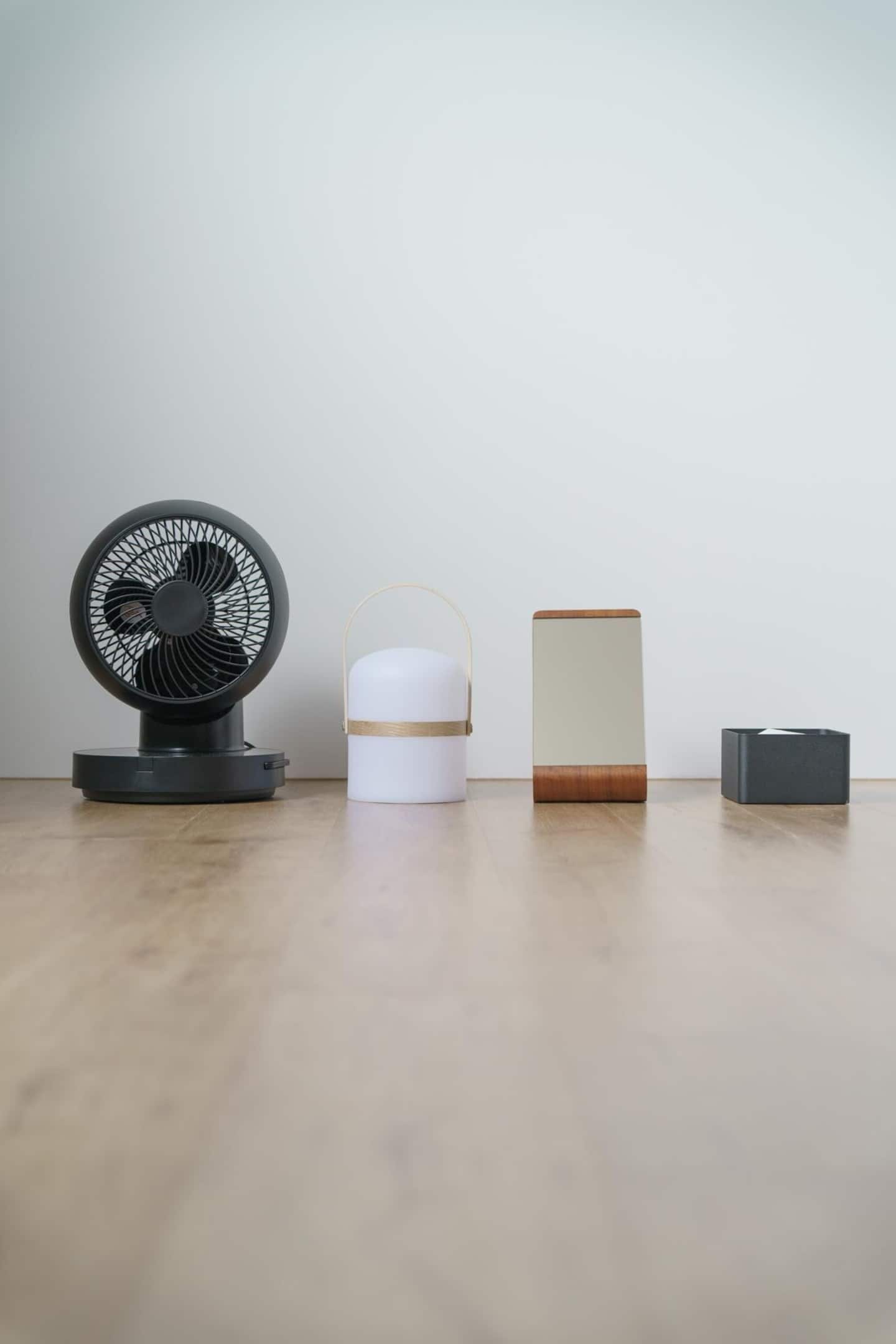 Um leque preto, um recipiente cilíndrico branco com cabo, um recipiente retangular com tampa de madeira e uma pequena caixa cinza sobre um piso de madeira em frente a uma parede lisa.