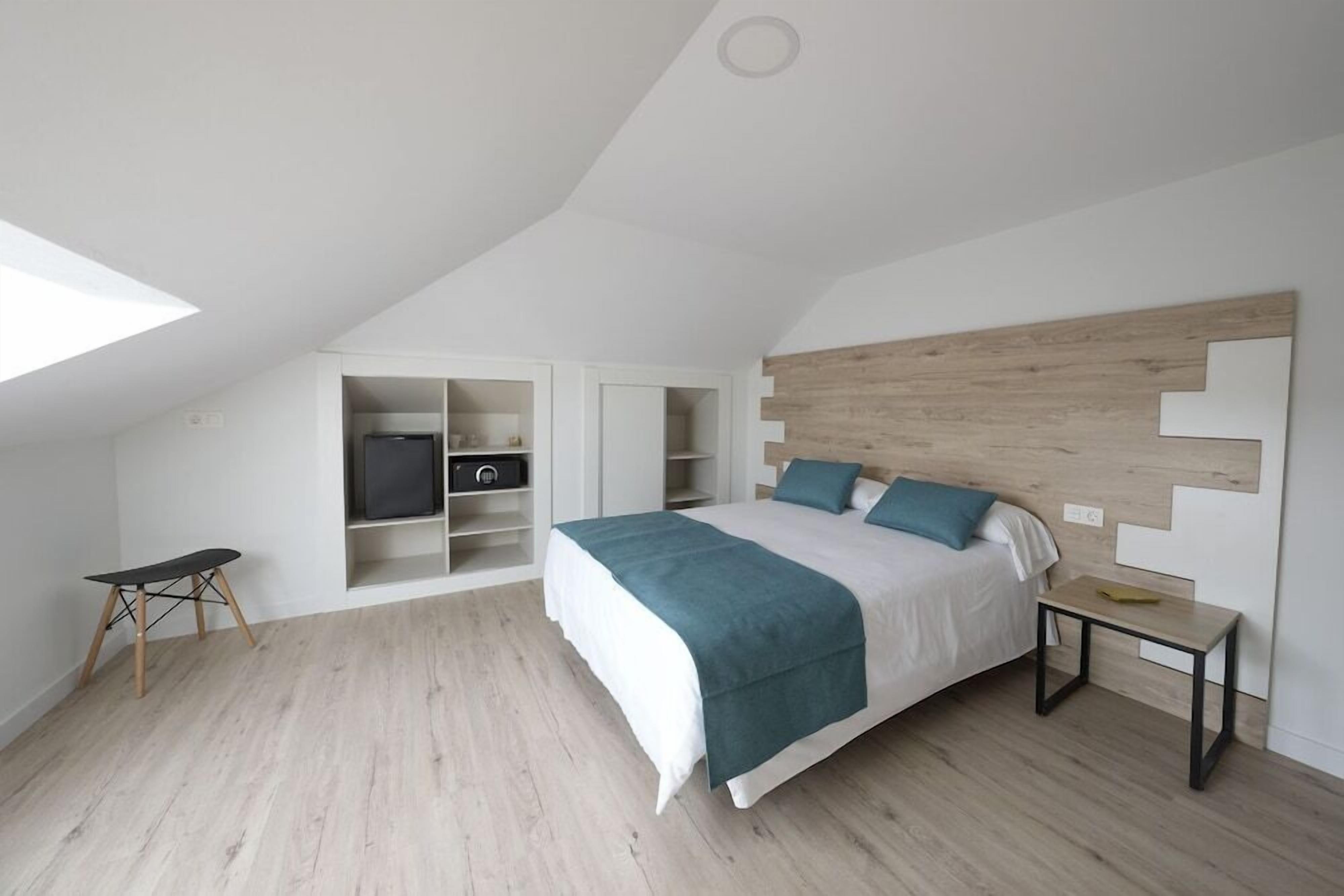 Quarto com teto inclinado, cama com cabeceira de madeira, mesa de cabeceira e prateleiras embutidas.
