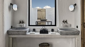 Casa de banho com duas pias de pedra, uma janela com vista para uma porta de madeira e nuvens, e espelhos fixados na parede.