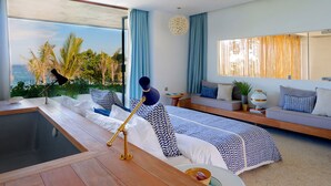 Quarto moderno com grande janela, piso de madeira, cama com roupa de cama estampada, mesa de cabeceira e vista para o oceano.