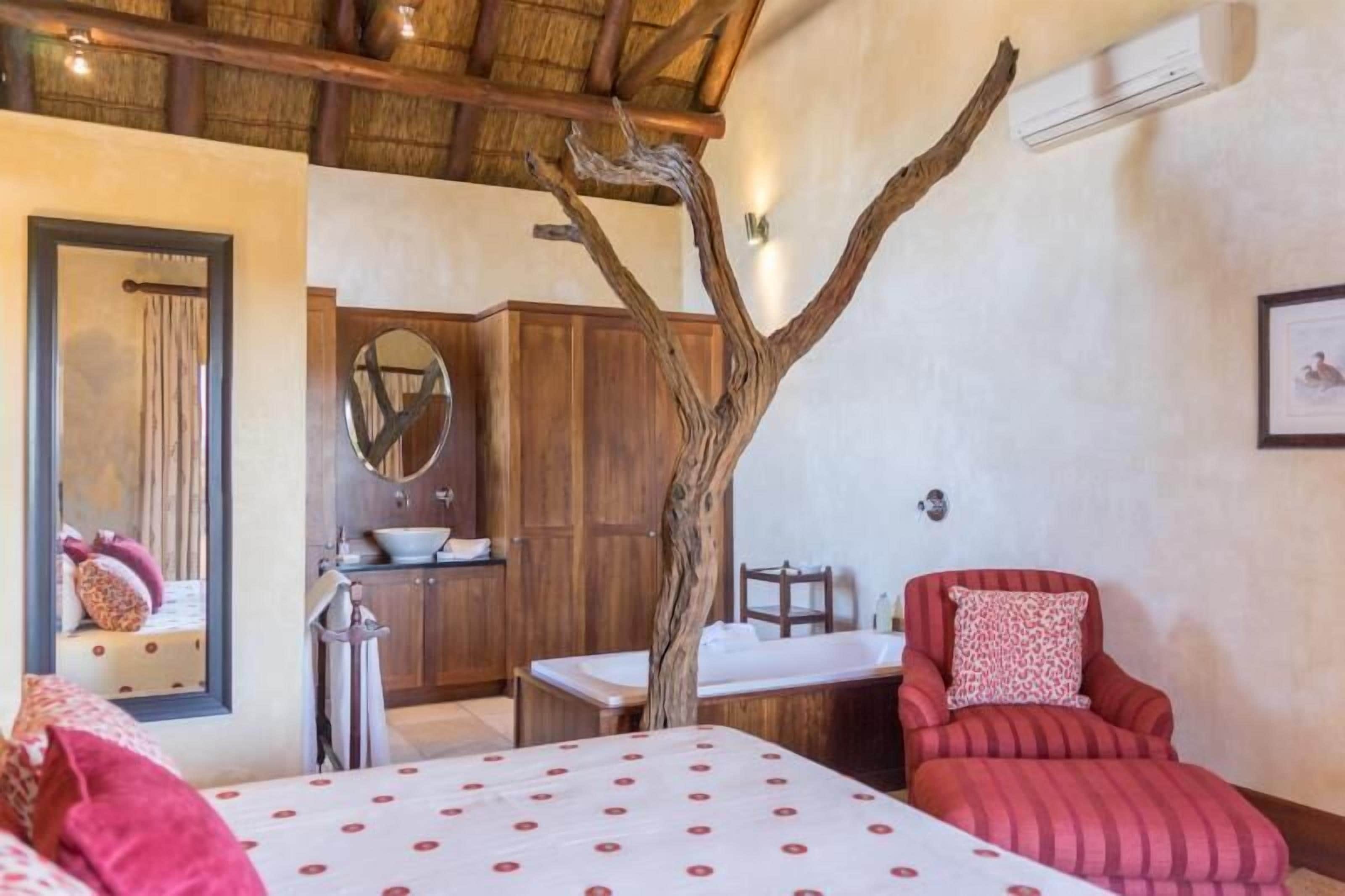 Quarto com cama, poltrona vermelha, escultura de árvore de madeira, banheiro com pia e espelho, e área de jantar com mesa e cadeiras.