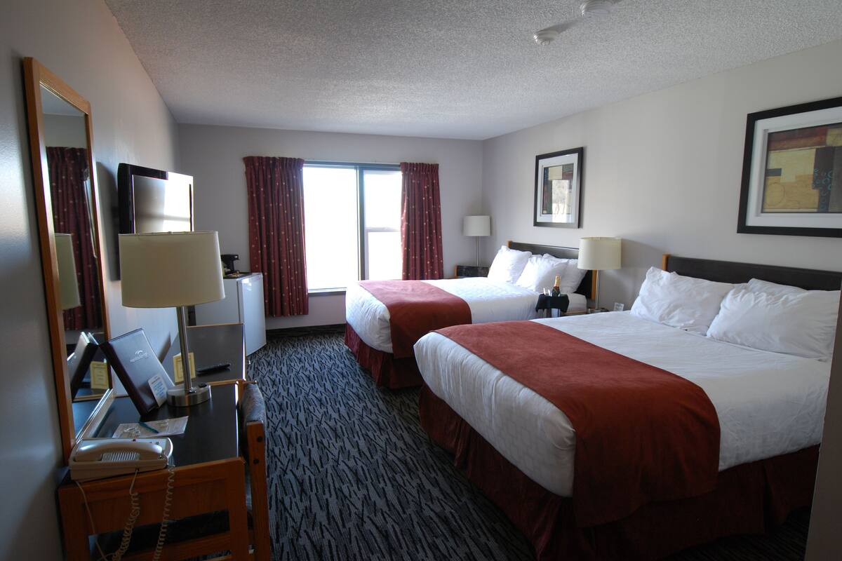 Quarto de hotel com duas camas, uma escrivaninha, um telefone, um espelho e uma janela com cortinas.