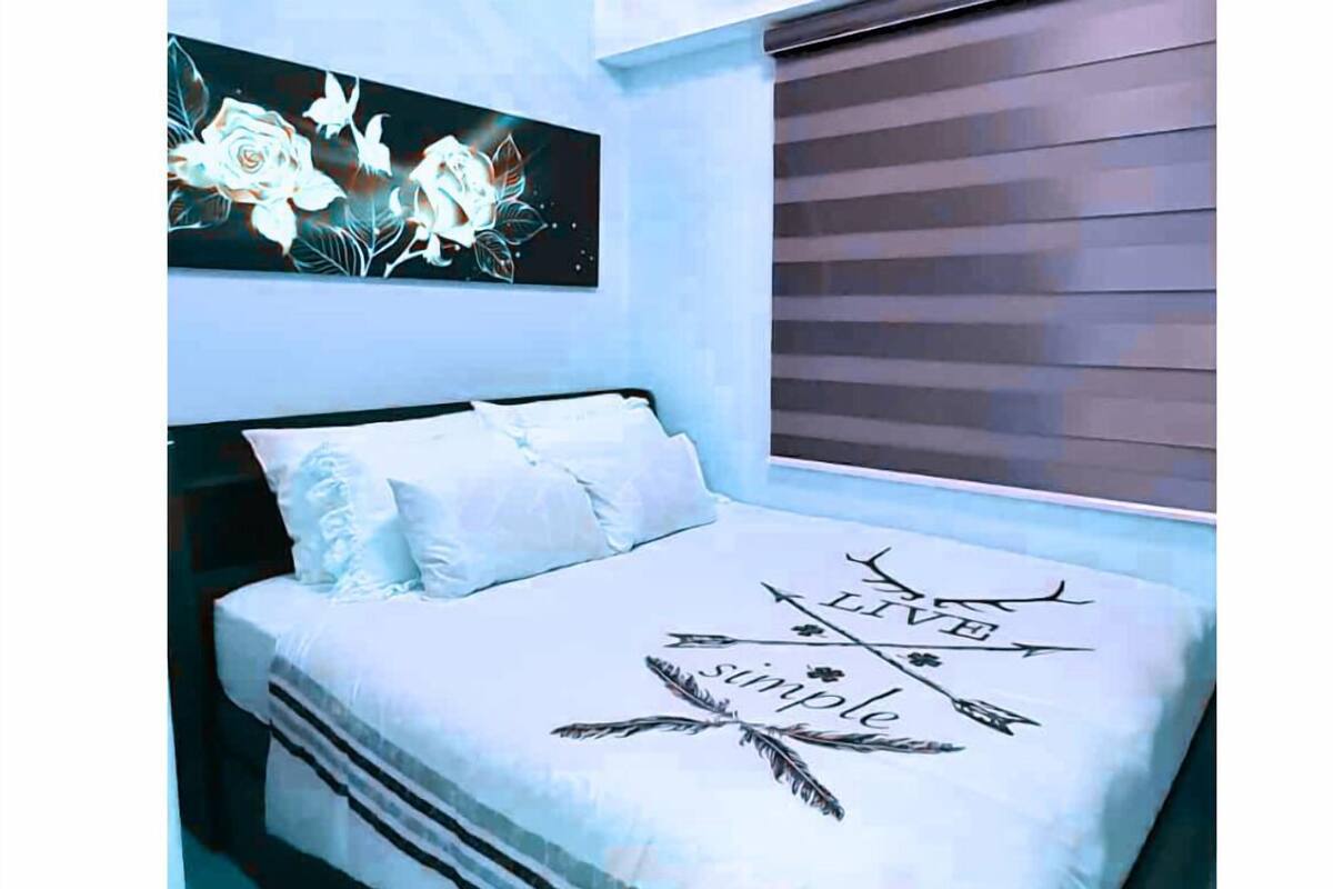 Quarto com cama, almofadas, obra de arte na parede e janela com persianas.