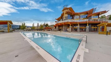 Uma piscina com placa de 'NÃO MERGULHAR', rodeada por um edifício de arquitetura moderna, com uma varanda notável e vista para as montanhas ao fundo.