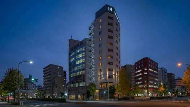 Um edifício de vários andares com uma entrada de esquina destacada, uma escadaria visível e fachada plana com janelas igualmente espaçadas.