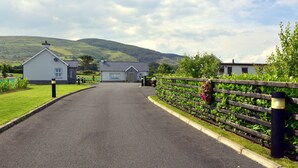 Uma estrada rural serpenteia até uma fileira de casas, com cerca de madeira e vegetação nas laterais.