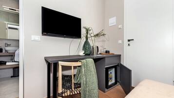 Quarto moderno com cama, escrivaninha com televisão, mini-frigorífico e banheiro ao fundo.