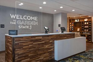 Balcão de recepção com a placa 'WELCOME TO THE GARDEN STATE', uma placa 'Gerente de Plantão' e a placa 'Hilton Honors'.