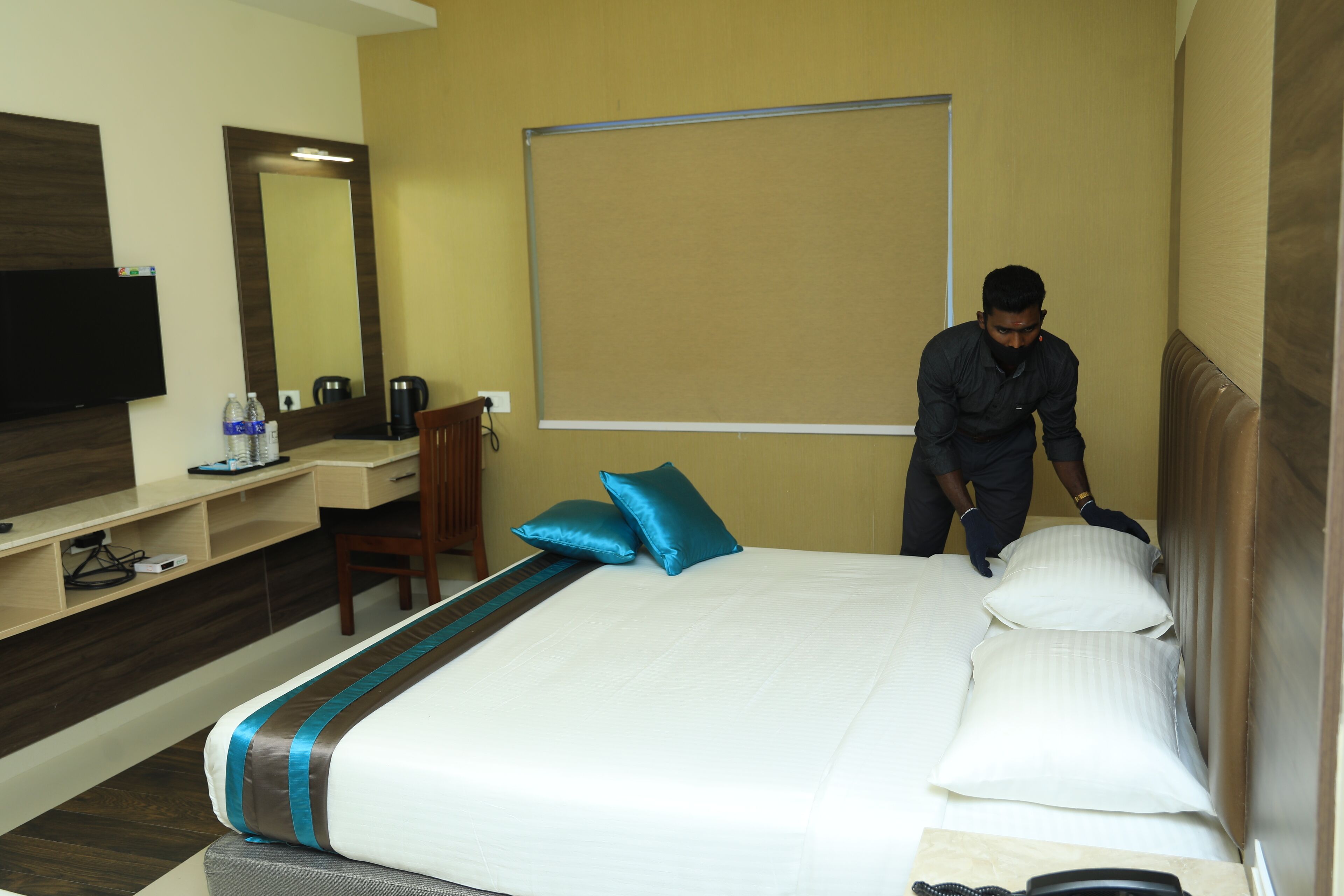 Quarto de hotel com cama, escrivaninha, cadeira, televisão e um espelho grande.