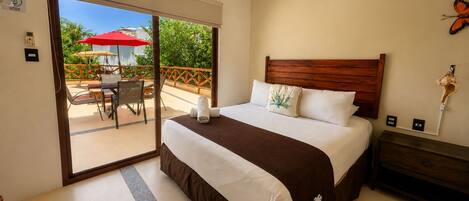 Quarto de hotel com uma cama, cabeceira de madeira, mesa de cabeceira, escrivaninha com cadeira e porta de vidro corrediça que dá para uma área externa com mesa e cadeiras.