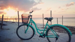 Uma bicicleta azul-turquesa com cesto numa praia arenosa ao pôr do sol.