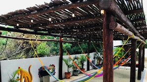 Área externa coberta com pérgola de madeira, redes coloridas e plantas em vasos.