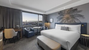 Quarto de hotel com uma cama grande, uma escrivaninha, uma cadeira, uma mesa pequena e vista para a cidade através de uma janela ampla.