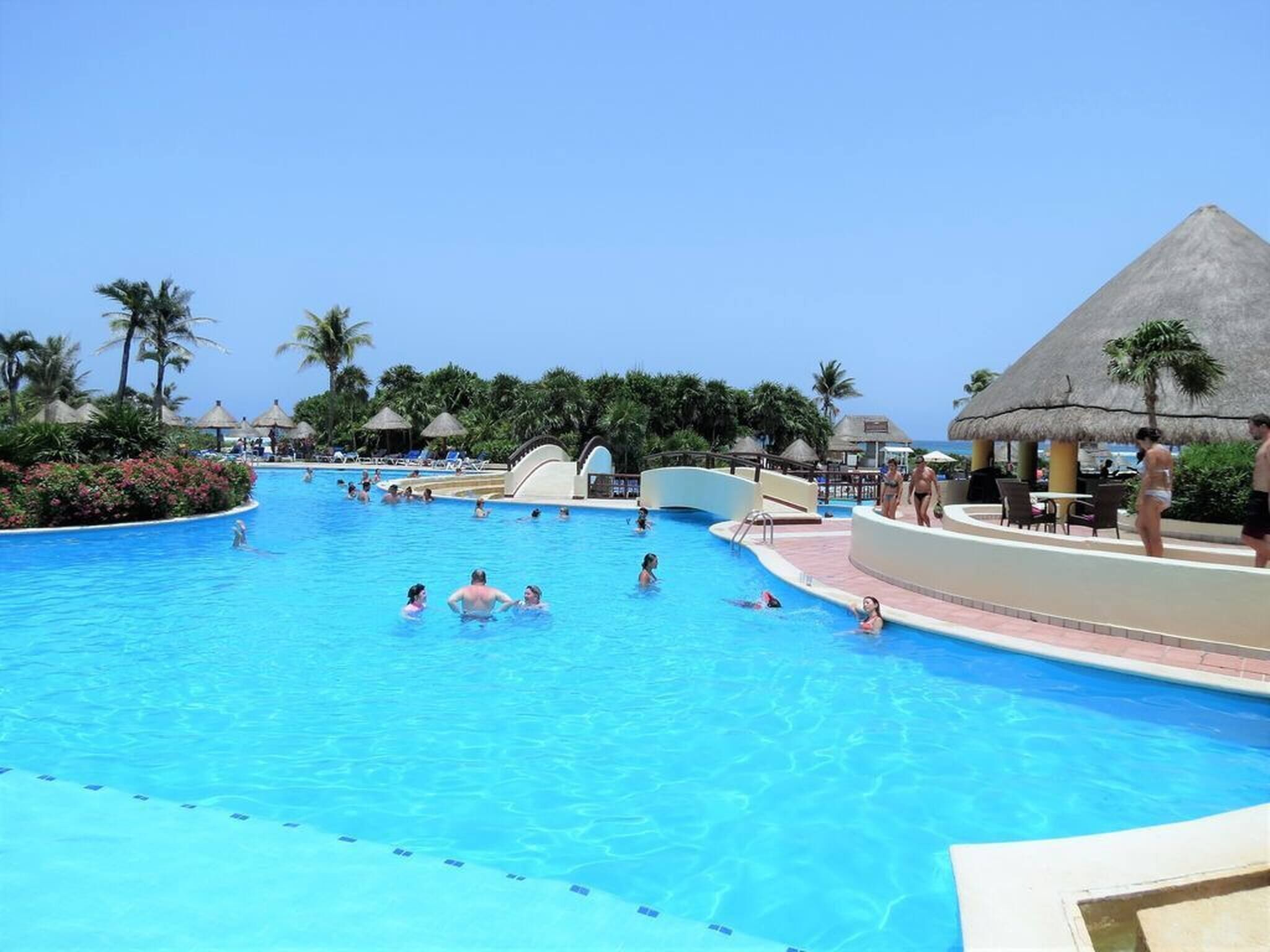 Uma piscina grande com várias pistas, rodeada por palmeiras e estruturas com telhados de palha.