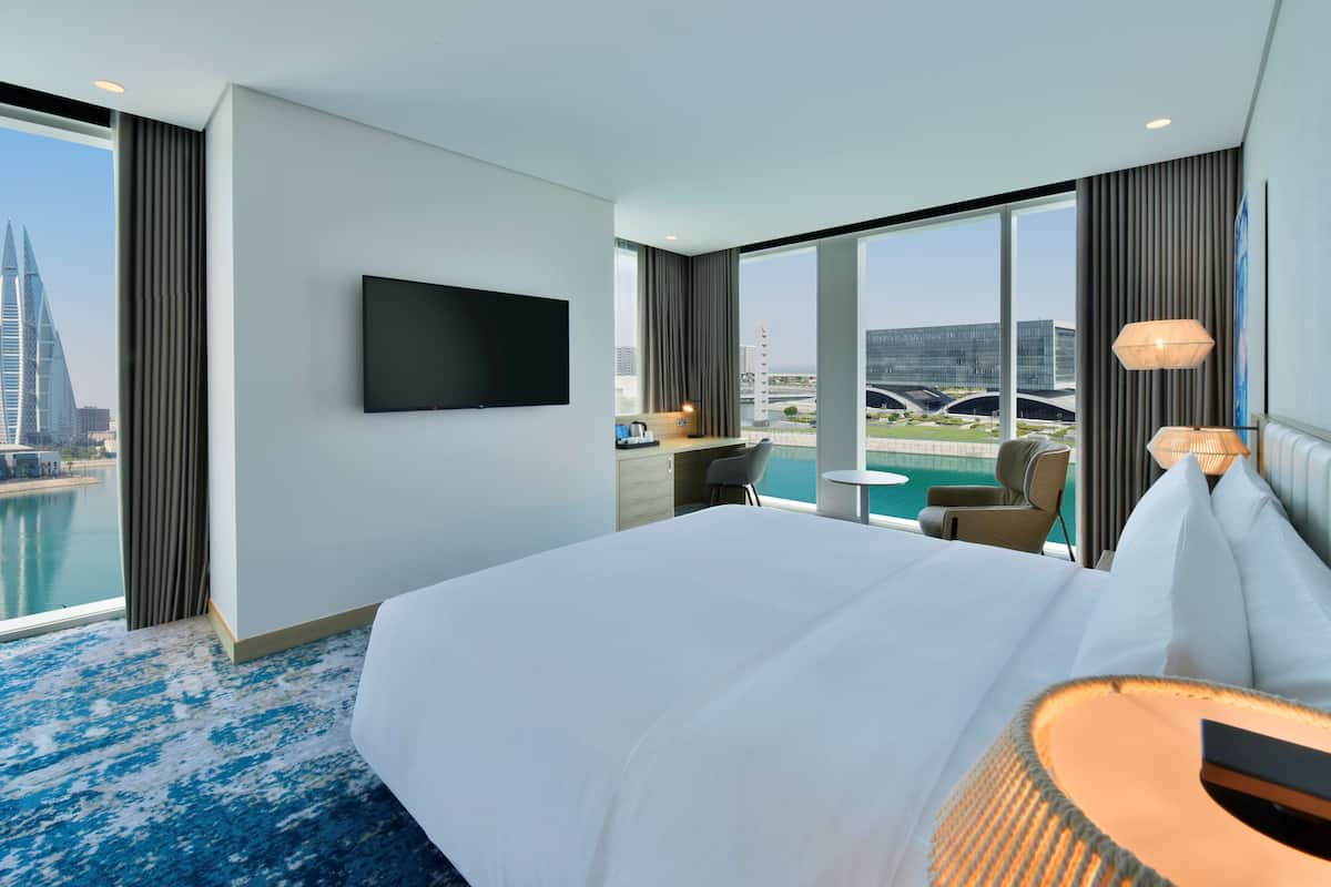 Quarto de hotel com uma cama grande, televisão de tela plana na parede, escrivaninha com cadeira e vista para a paisagem urbana e um corpo de água através de amplas janelas.
