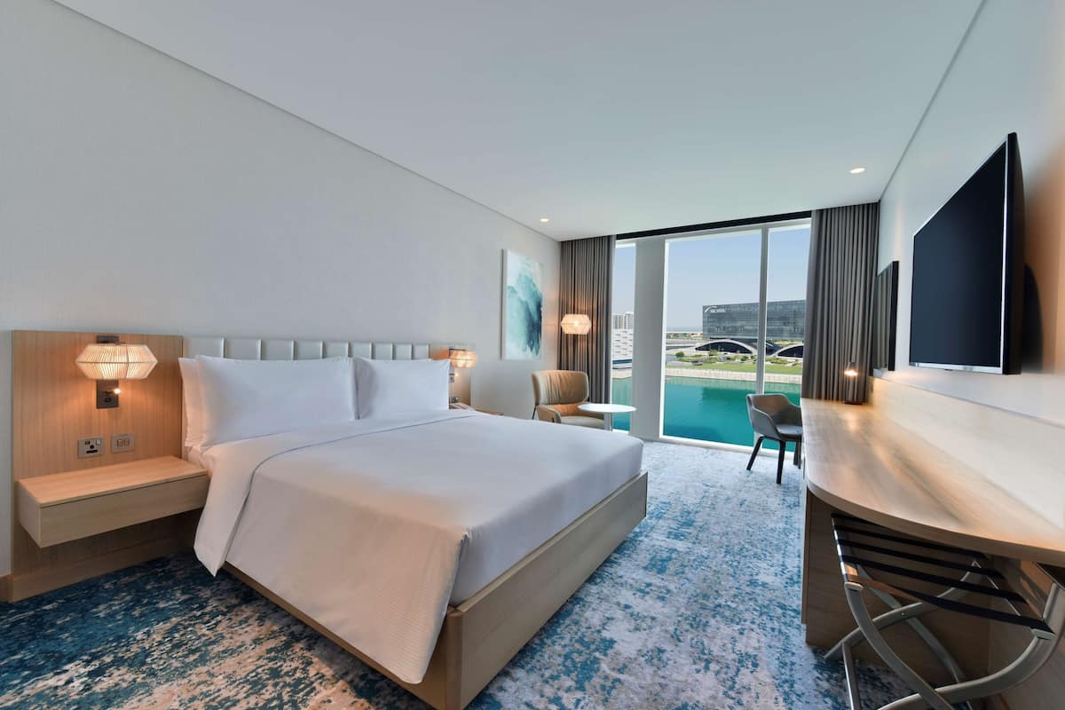 Quarto de hotel moderno com uma cama grande, uma escrivaninha com cadeira, TV e vista para uma piscina.