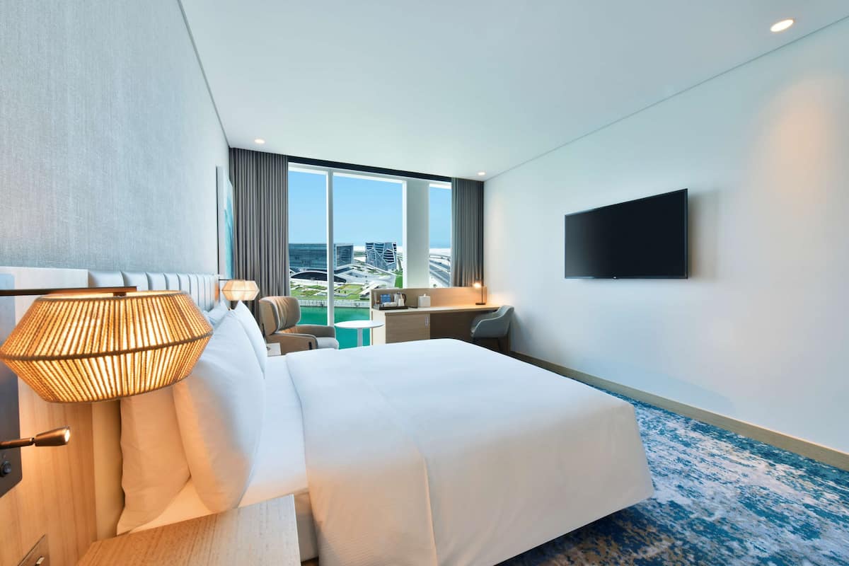 Quarto de hotel com uma cama grande, uma escrivaninha, uma cadeira, uma televisão e vista para a paisagem urbana.