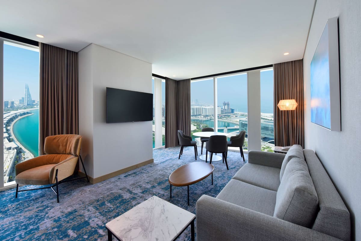 Quarto de hotel moderno com uma janela grande, um sofá, uma cadeira, uma mesa pequena e uma televisão de tela plana.