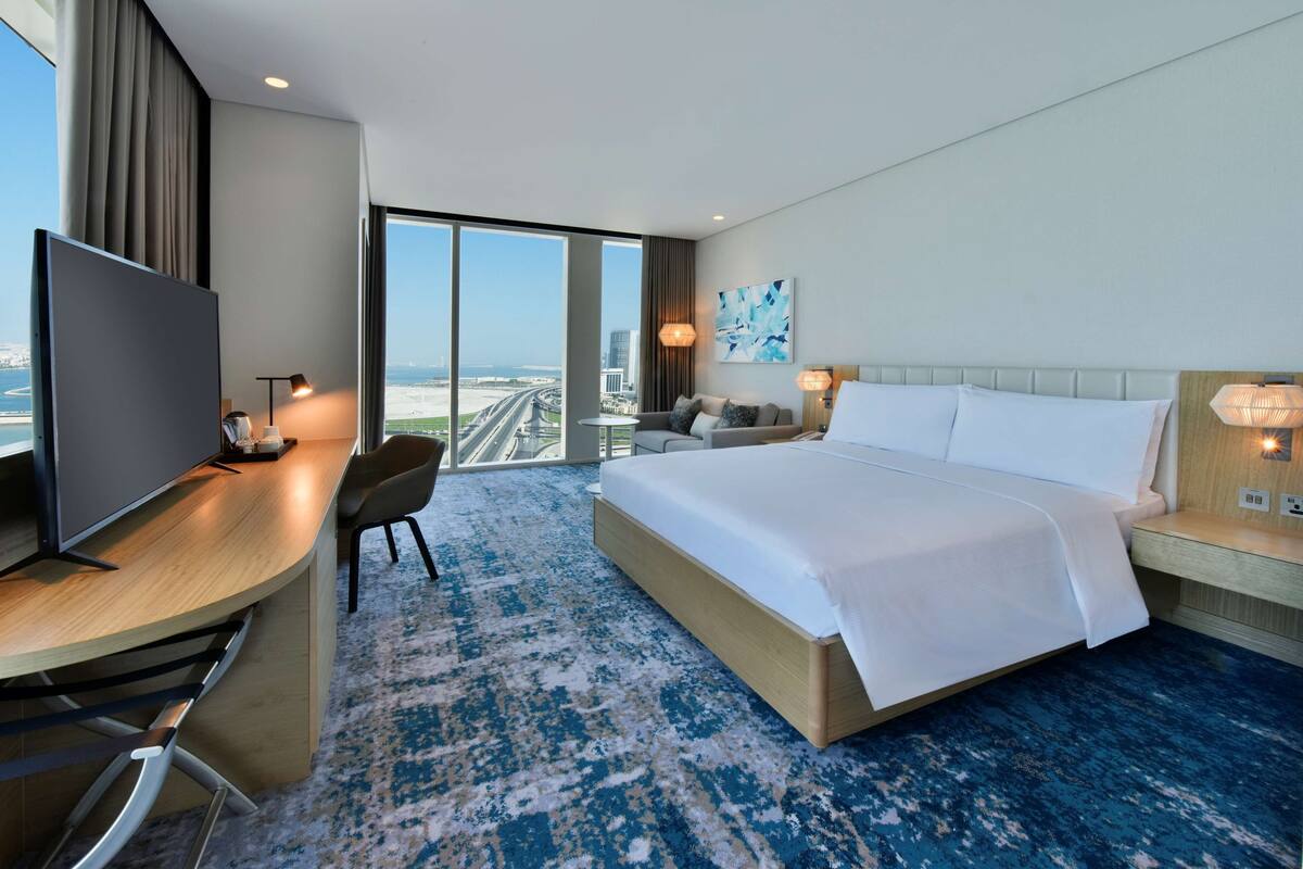 Quarto de hotel com uma cama grande, uma mesa de trabalho com uma TV, uma cadeira e vista para a paisagem urbana.
