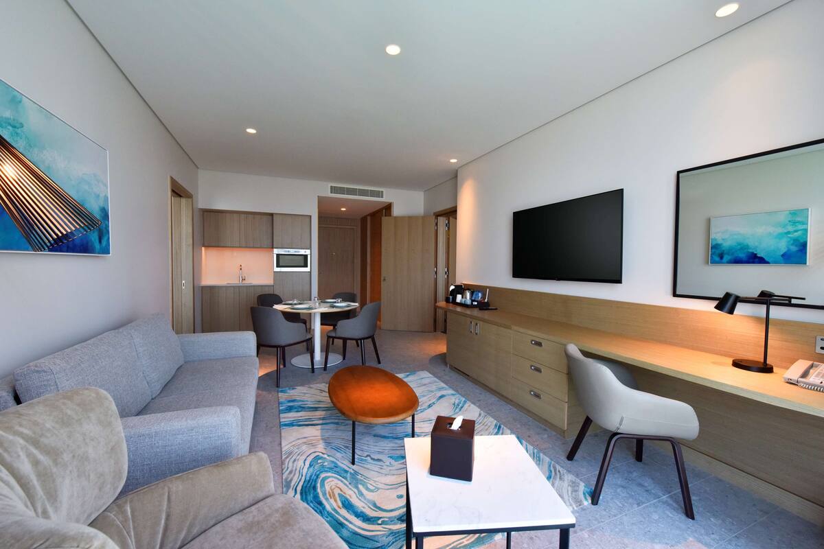 Quarto de hotel moderno com sofá, mesa de jantar, área de cozinha e uma televisão grande na parede.