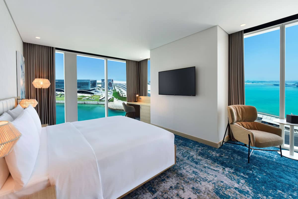 Quarto de hotel com uma cama grande, uma escrivaninha, uma cadeira, uma televisão e vista para o oceano e uma ponte.