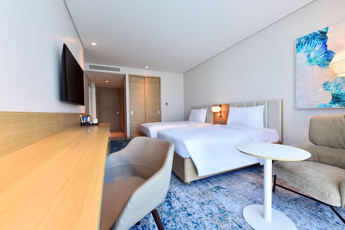 Quarto de hotel com cama, duas mesas de cabeceira, uma cadeira, escrivaninha e televisão.