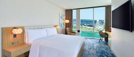 Quarto de hotel com uma cama grande, uma mesa, uma cadeira, uma televisão e vista para uma piscina e edifícios.