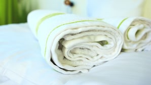 Toalha branca dobrada com uma faixa verde, colocada sobre uma cama com roupa de cama branca.