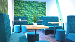 Uma área de estar moderna com assentos estofados em tom de verde-água, uma mesa de madeira e um mural de parede com folhagens verdes.