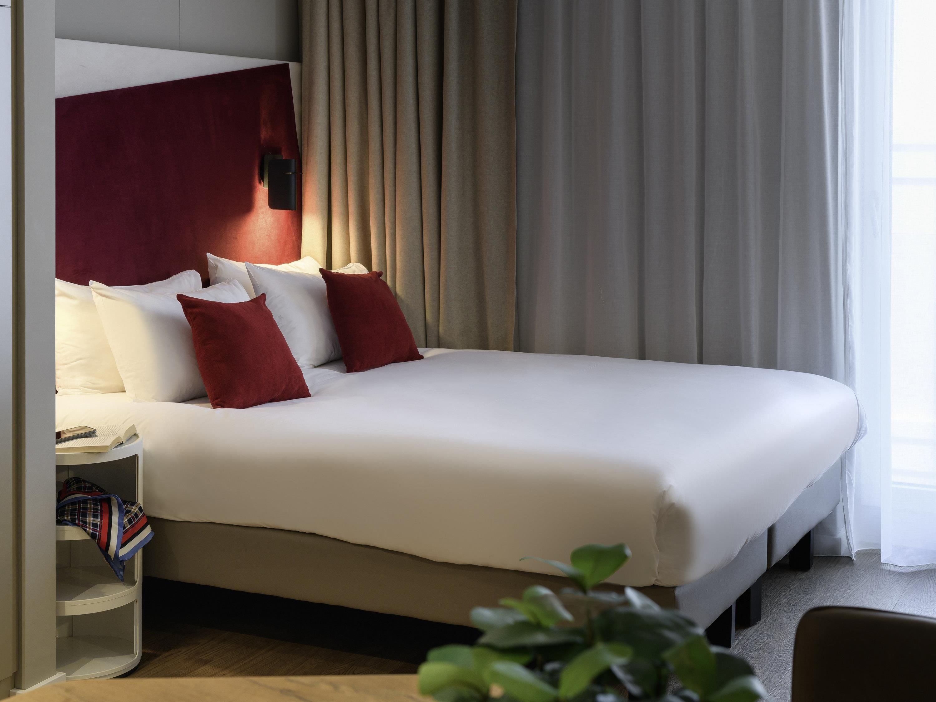 Cama bem arrumada com travesseiros brancos e vermelhos, mesa de cabeceira com livros e uma planta em primeiro plano.