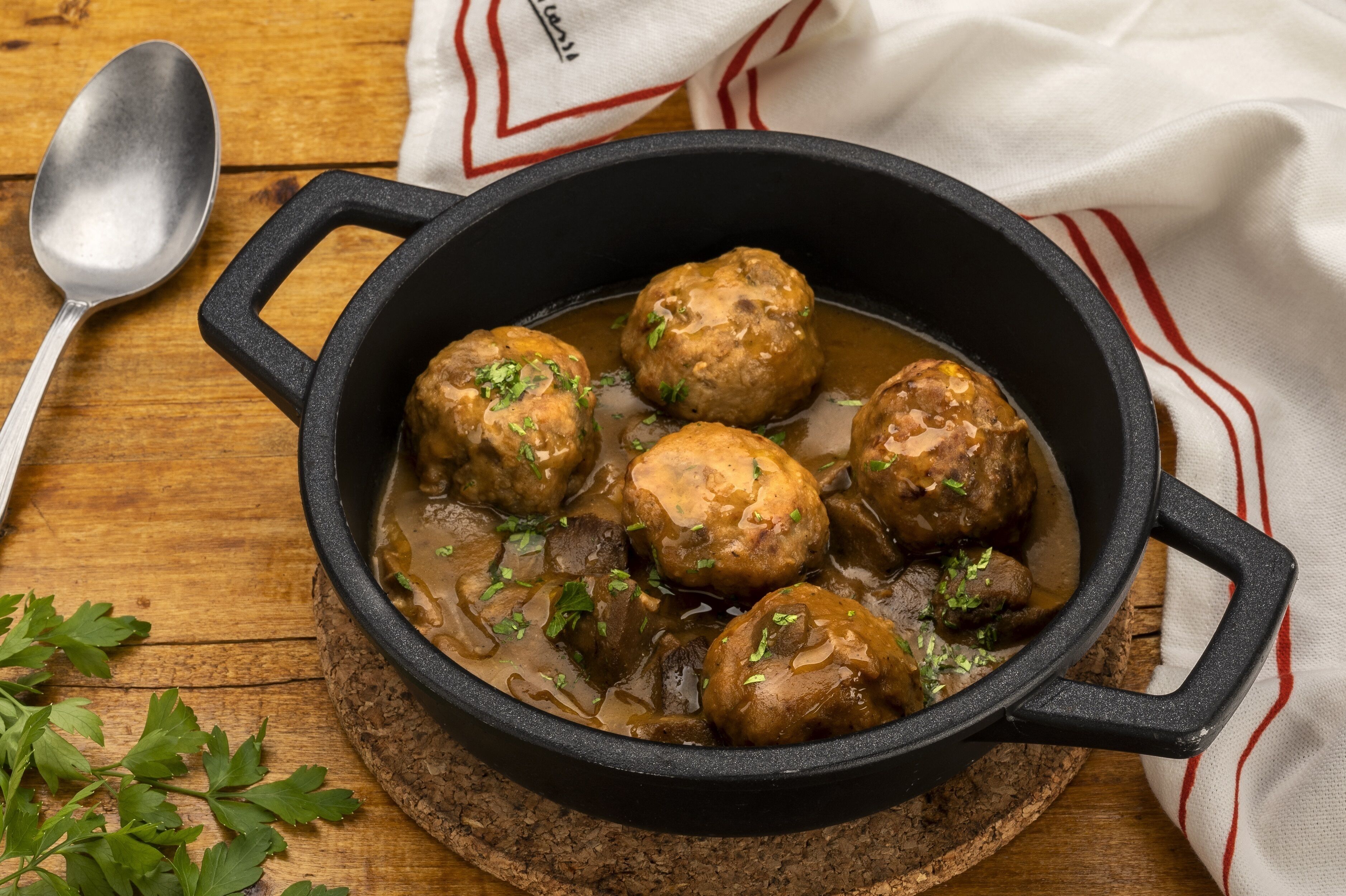 Uma frigideira de ferro fundido com almôndegas ao molho, colocada sobre uma superfície de madeira, com uma colher e folhas de salsa por perto.