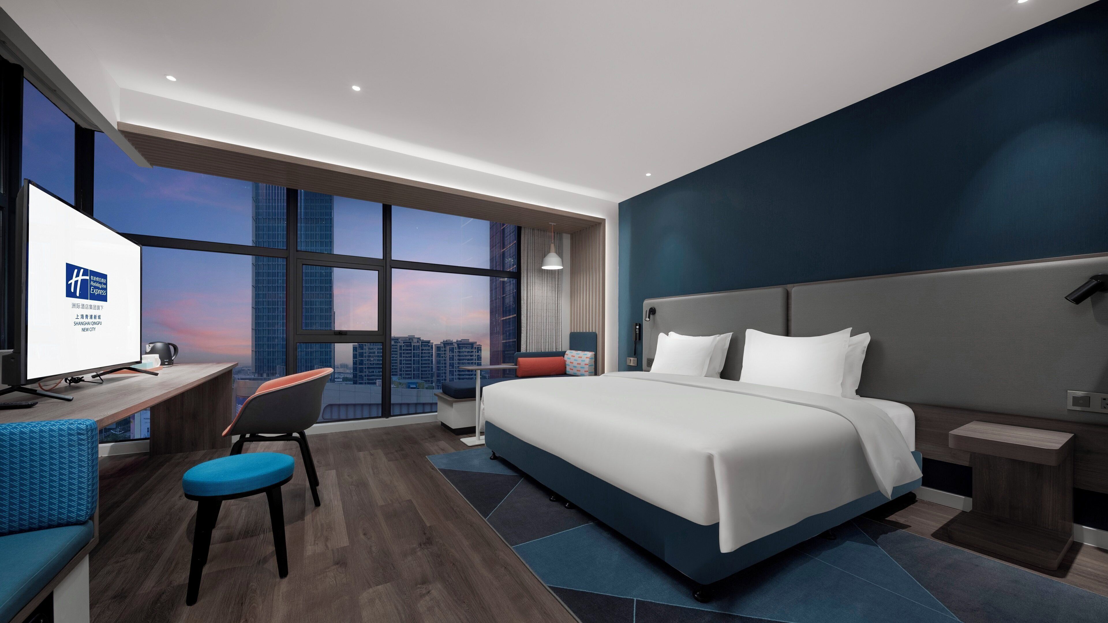 Quarto de hotel moderno com uma cama grande, escrivaninha com computador, cadeira e vista da linha do horizonte da cidade por grandes janelas.