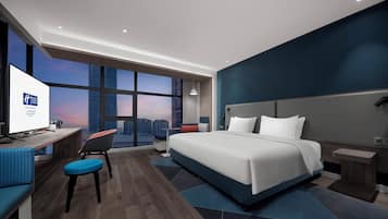 Quarto de hotel moderno com uma cama grande, escrivaninha com computador, cadeira e vista da linha do horizonte da cidade por grandes janelas.