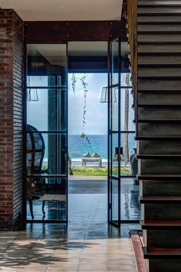 Uma porta de vidro aberta dá para um alpendre com vista para o mar, um banco e uma cadeira.