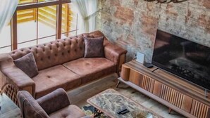 Uma sala de estar acolhedora com um amplo sofá de couro, uma mesa de centro e uma televisão de tela plana. O espaço possui vigas de madeira aparente, uma parede de tijolos e uma grande janela com cortinas leves.