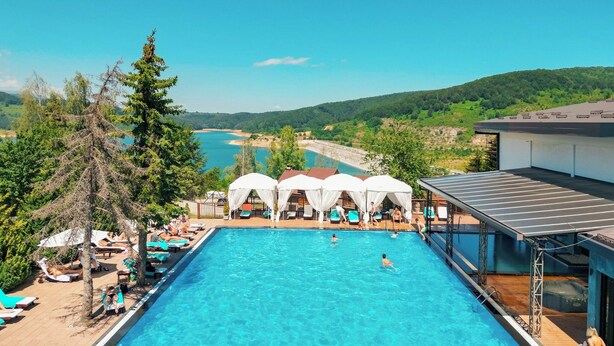 Piscina com água azul cristalina, rodeada por espreguiçadeiras e guarda-sóis, com vista para um morro arborizado e um corpo de água ao fundo.