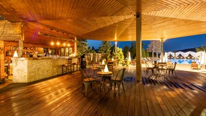 Um deck de madeira com área de estar externa, bar e piscina ao fundo.