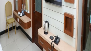 Quarto de hotel com um espelho grande, televisão, armário de madeira, mesa pequena com chaleira e telefone, e uma cadeira.