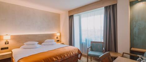 Quarto de hotel com uma cama grande, duas luminárias laterais, uma cabeceira de madeira, uma janela com cortinas, uma cadeira e uma mesa com bandeja.