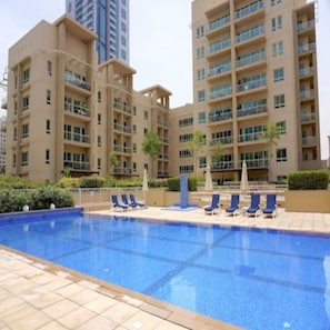 Piscina com espreguiçadeiras e um edifício com vários andares e varandas ao fundo.