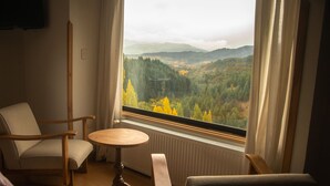 Quarto com piso de madeira, duas poltronas, uma mesa redonda pequena e uma janela com vista para uma paisagem arborizada.