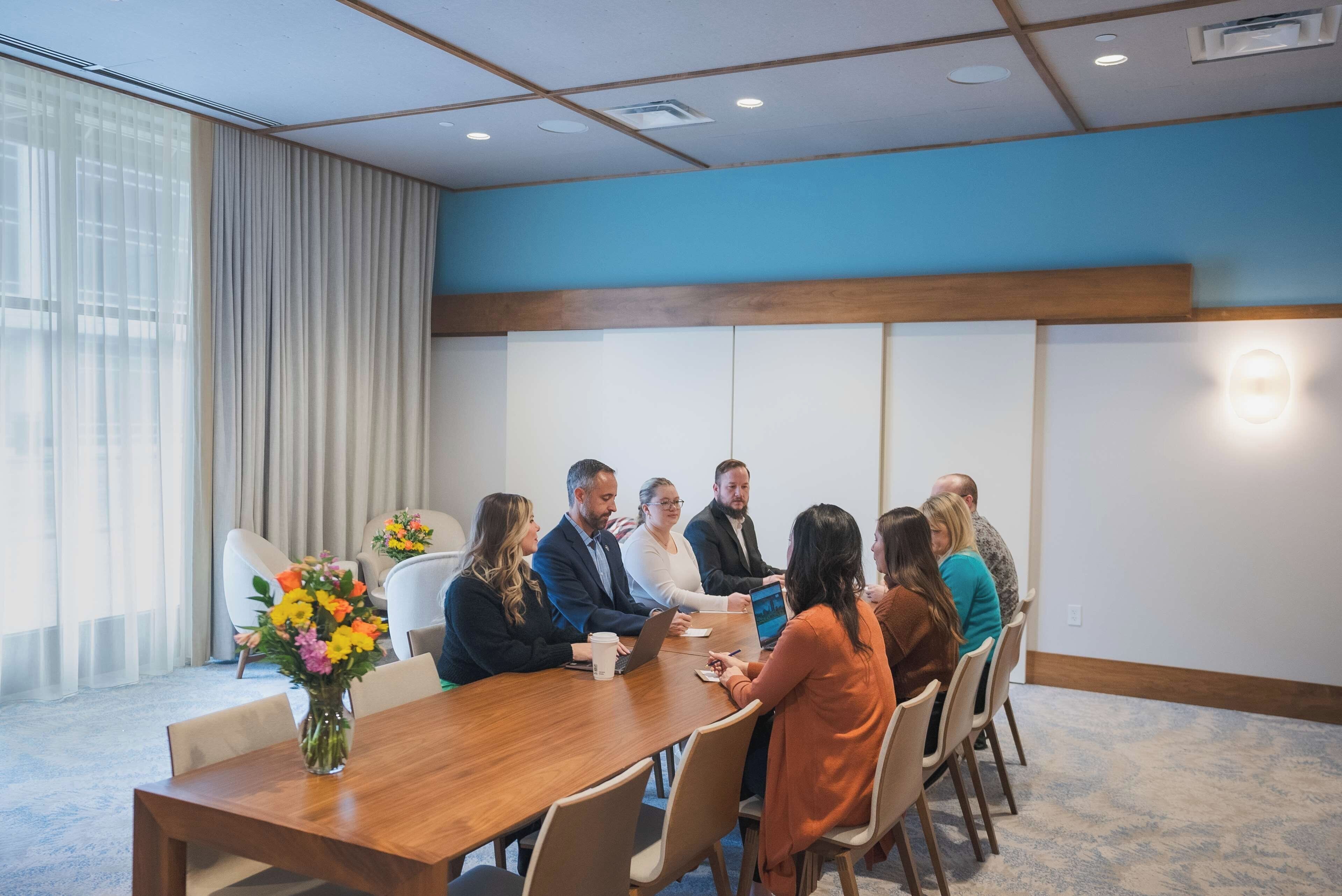 Uma sala de conferências com uma mesa comprida, cadeiras e um vaso de flores.