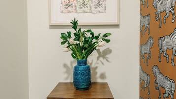 Uma obra de arte emoldurada na parede, um vaso azul com flores brancas sobre uma mesinha de madeira e um papel de parede estampado na parede vizinha.