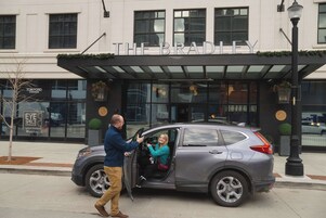 Um homem e uma mulher saindo de um carro cinzento em frente a um edifício com uma placa que diz 'THE BRADLEY'.