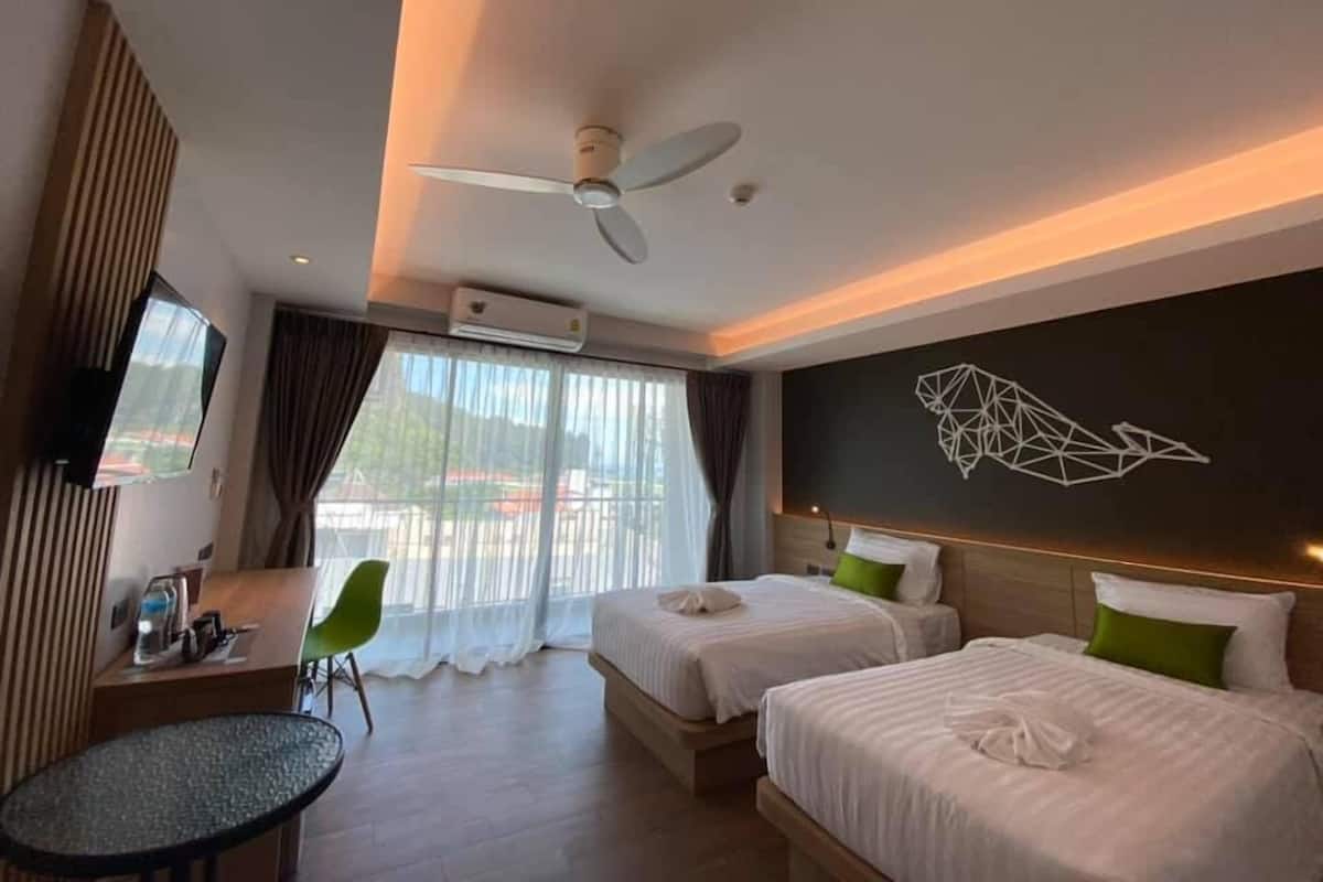 Quarto de hotel com duas camas, uma escrivaninha, uma cadeira e um ventilador de teto.
