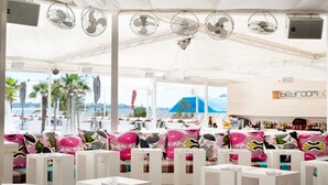 Restaurante à beira-mar com mesas brancas, almofadas coloridas, um bar e vista para o mar.