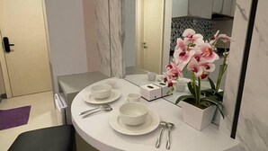 Uma área de jantar moderna com uma mesa redonda posta para dois, pratos, tigelas e talheres brancos. Há uma pequena orquídea em vaso sobre a mesa, e um espelho reflete o espaço.