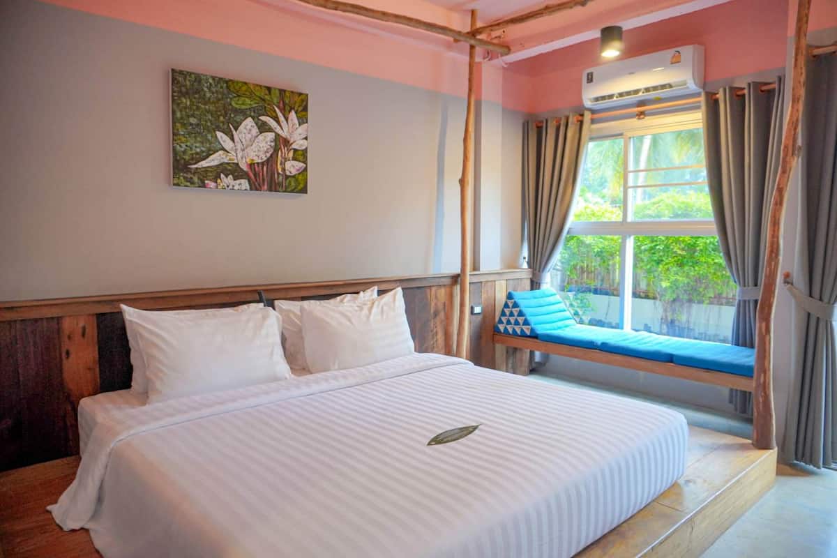 Quarto com cama, um assento junto à janela com almofada azul, ar condicionado e um quadro de flores na parede.