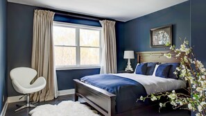 Quarto com cama, cadeira, mesa de cabeceira, abajur e janela com cortinas.