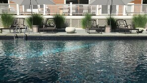 Piscina com deck branco, cadeiras de descanso e um grande vaso de plantas.